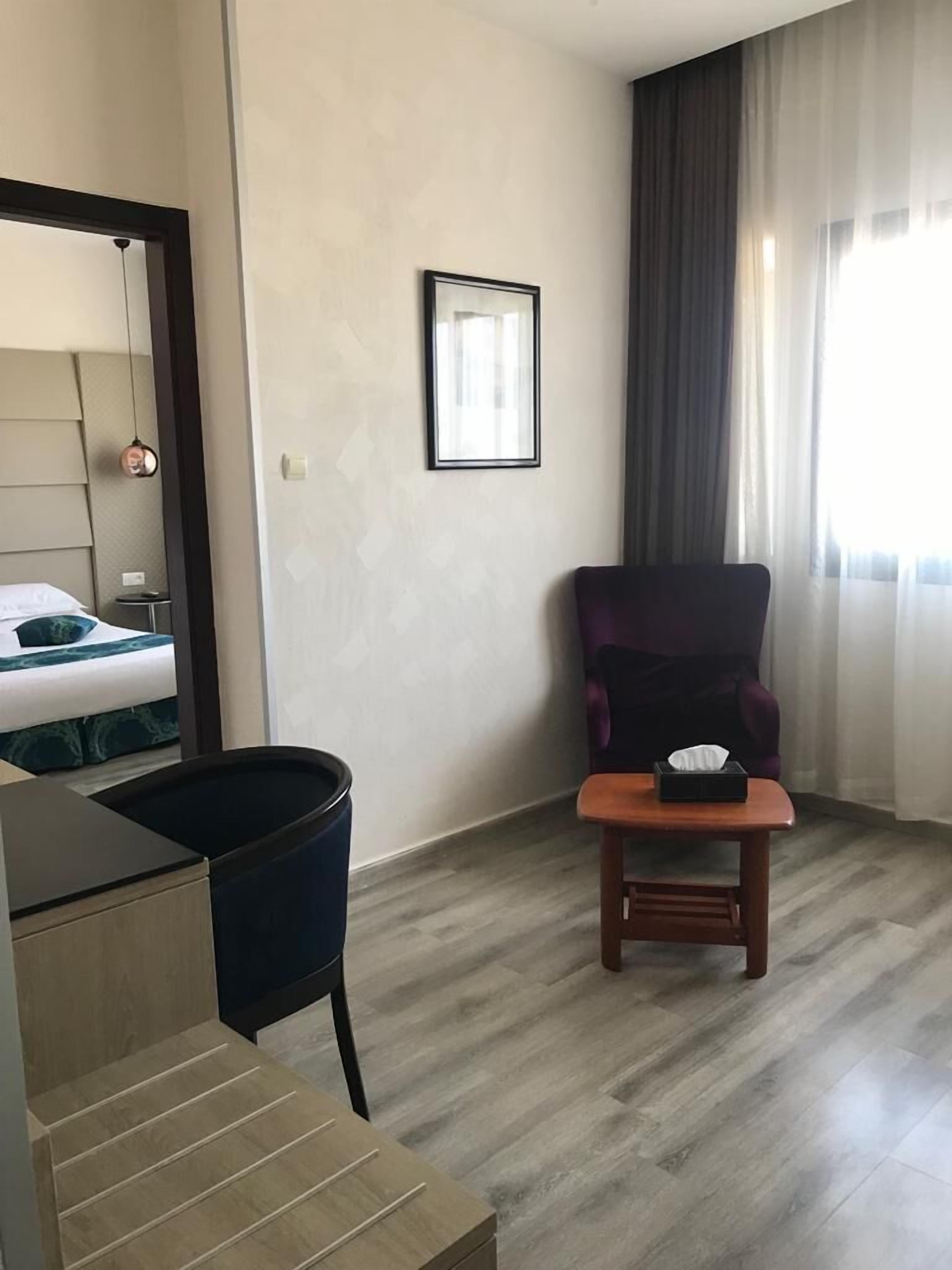 Quarto de hotel com cama, escrivaninha, cadeira, mesinha de centro com caixa de lenços, quadro na parede e janela com cortinas.