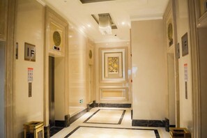 Corredor de hotel com piso de mármore, elevador e painel decorativo na parede.