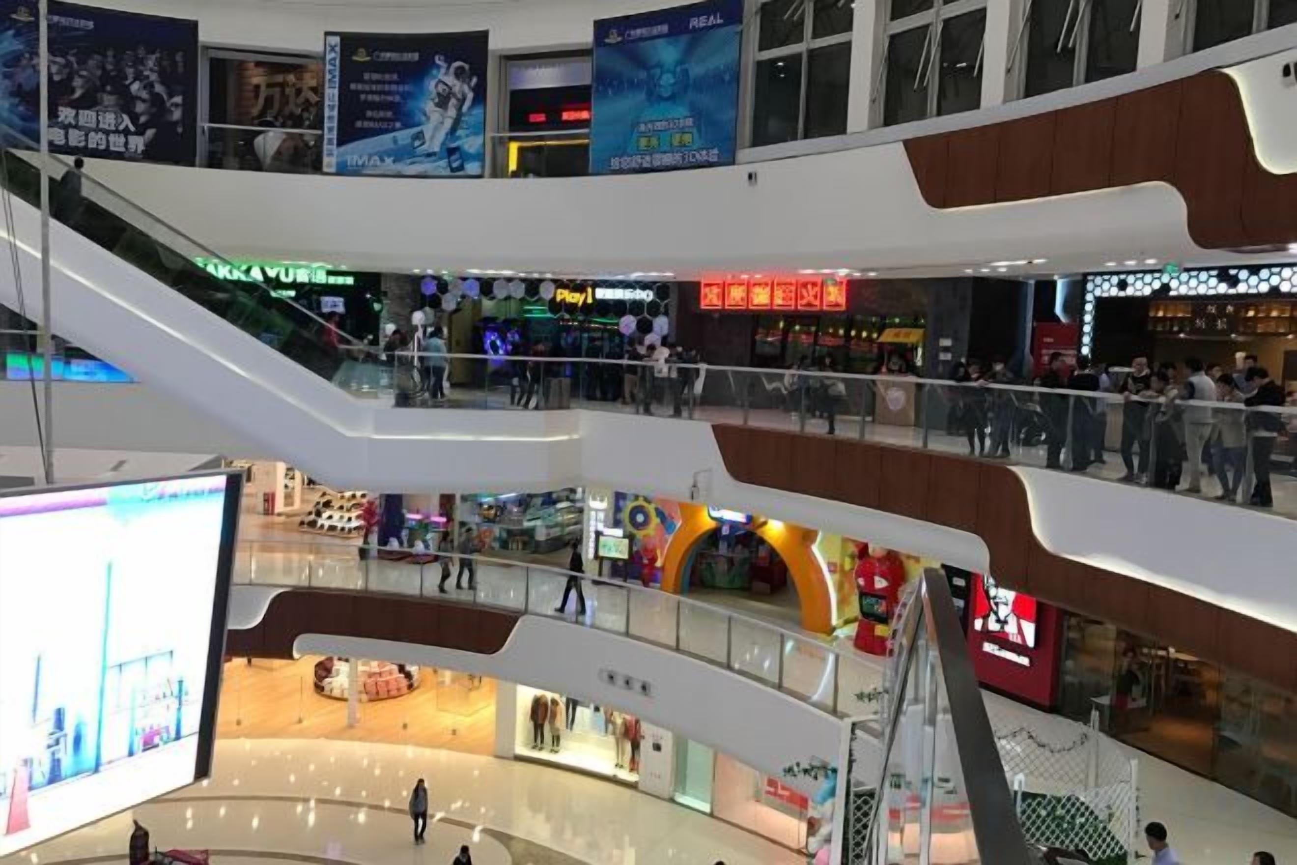 Um shopping center com vários níveis, escadas rolantes, lojas e uma grande tela de anúncios digitais.