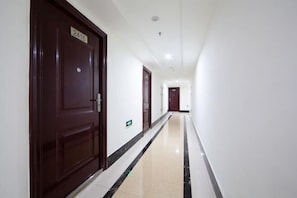 Um corredor comprido e estreito, com portas de madeira escura, paredes brancas e chão de mármore.