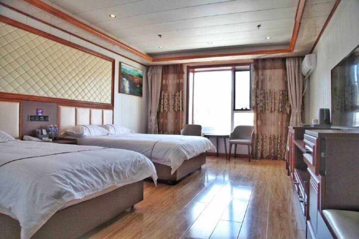 Quarto de hotel com duas camas, uma escrivaninha, uma cadeira, uma televisão e uma janela grande.
