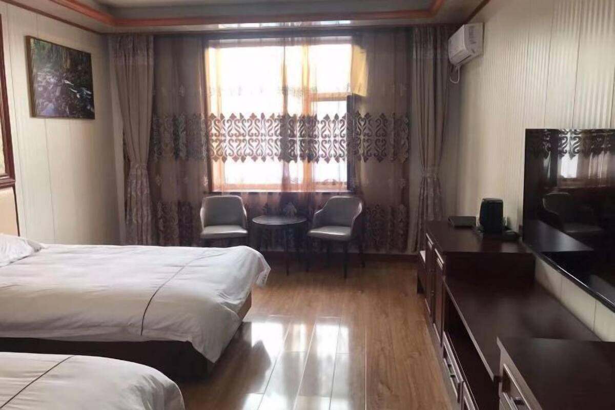 Quarto de hotel com duas camas, televisão, pequena mesa com cadeiras e janela com cortinas.