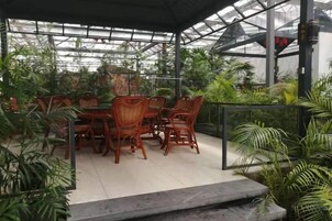 Pátio externo com móveis de vime e teto de vidro, cercado por diversas plantas.