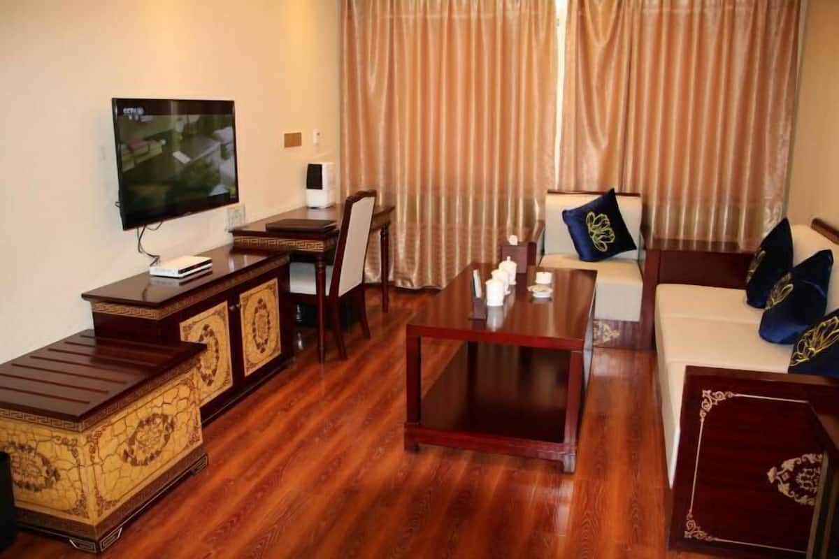 Quarto de hotel com piso de madeira, televisão sobre suporte, sofá com almofadas decorativas, mesa pequena e armário com detalhes elaborados.