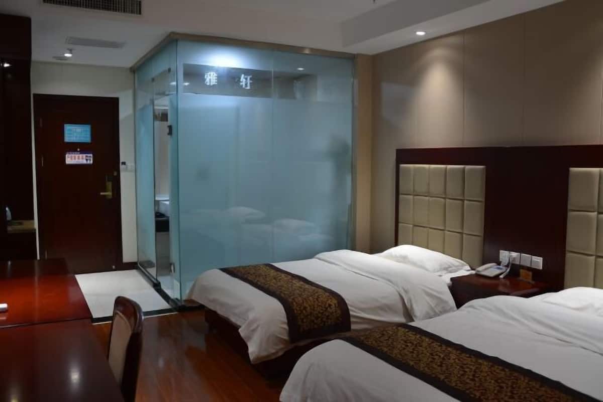 Quarto de hotel com duas camas, uma escrivaninha de madeira, uma divisória de vidro e uma porta escura.