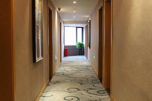 Um corredor de hotel com obras de arte emolduradas nas paredes, um tapete estampado e uma planta ao fundo.