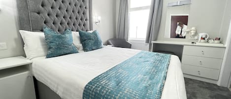 Quarto de hotel com uma cama grande, cabeceira estofada cinza, mesa de cabeceira branca, poltrona cinza, escrivaninha com espelho e janela com cortinas cinzas.