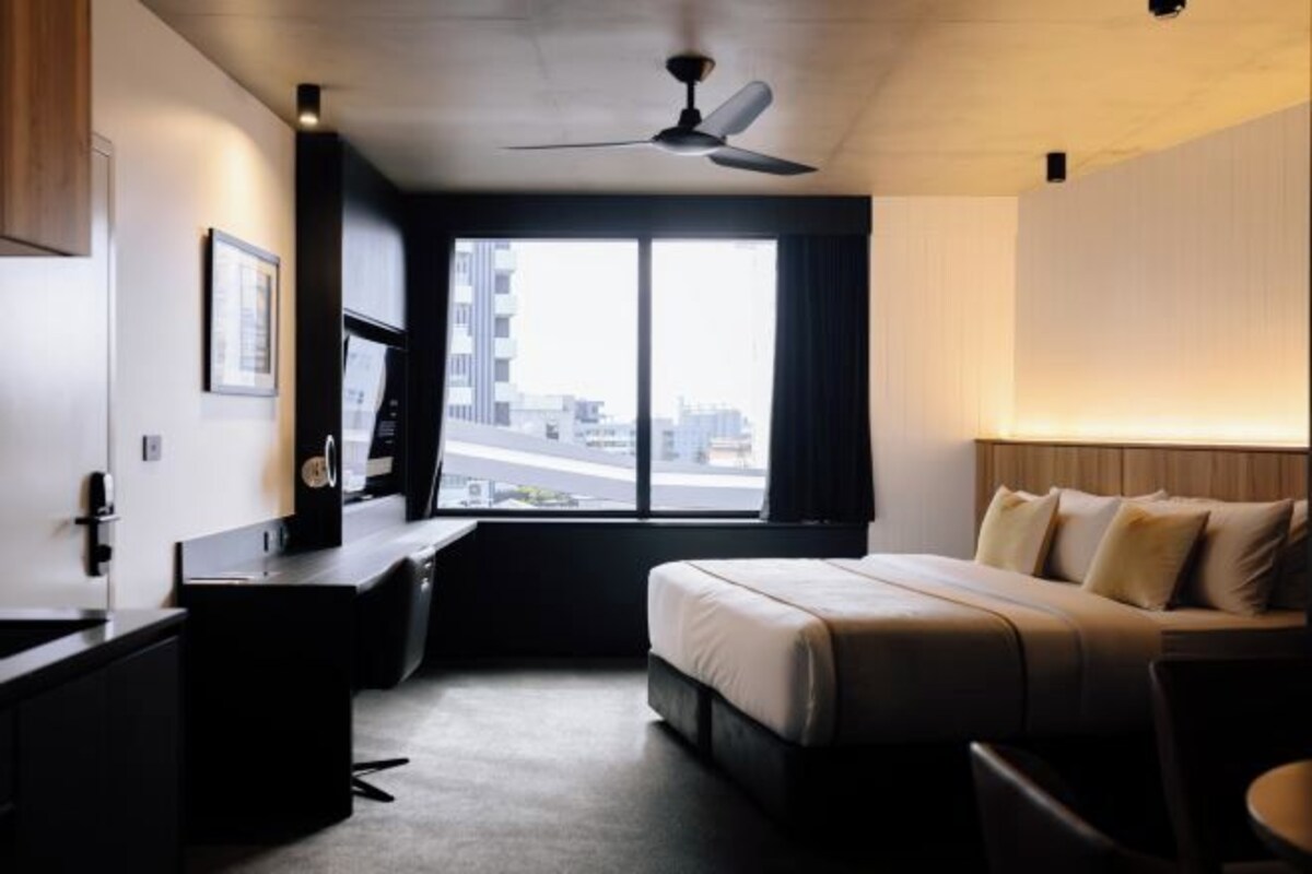 Quarto de hotel com cama, escrivaninha e uma janela grande com vista para a cidade.