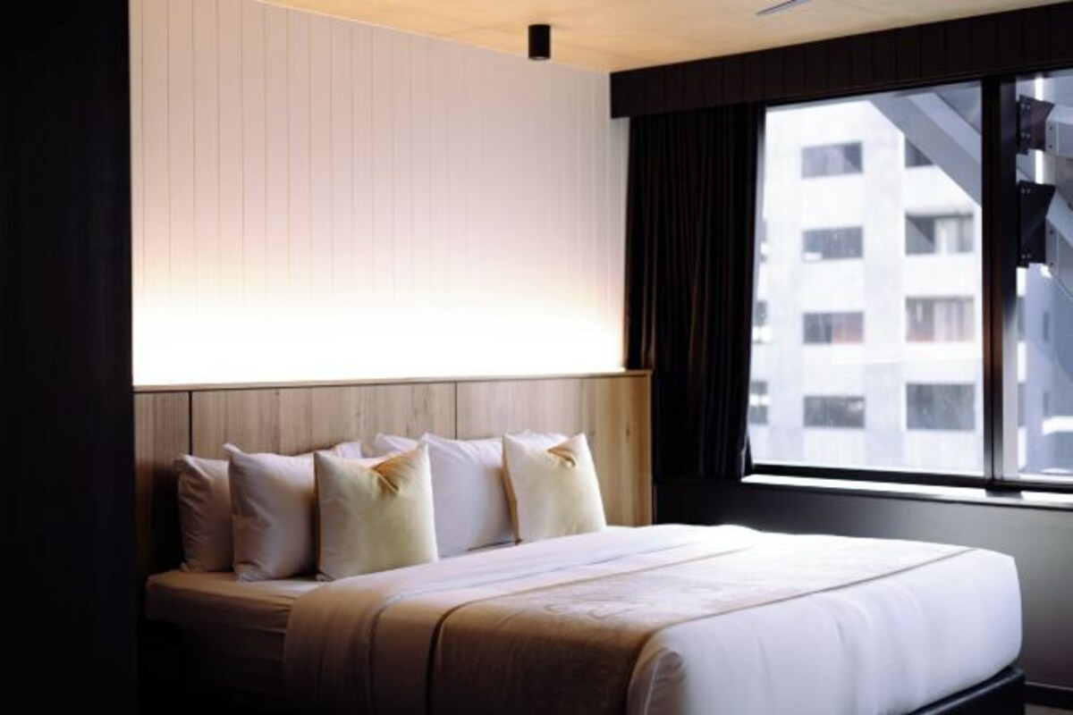 Quarto de hotel com uma cama grande, cabeceira com linhas verticais, janela com vista para edifícios e cortina escura.