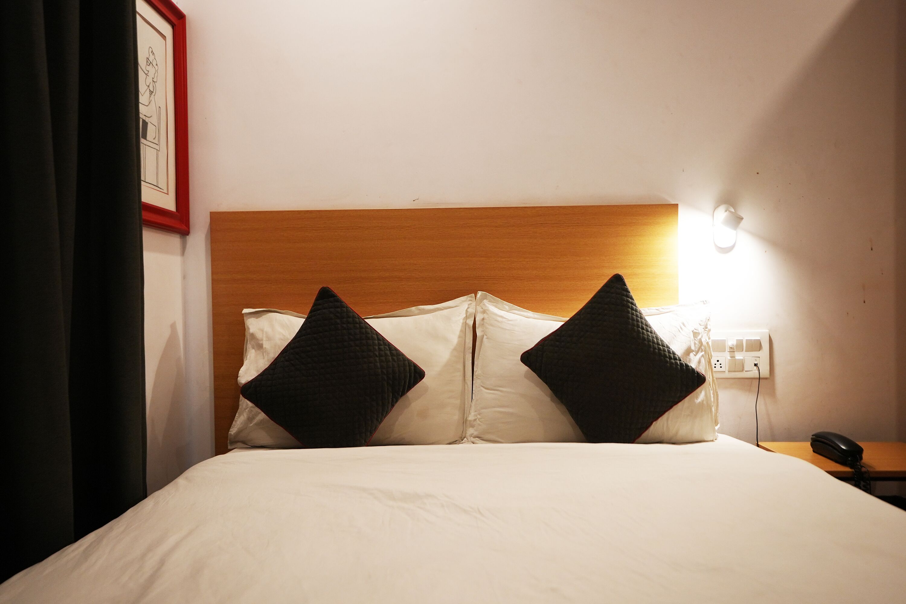 Quarto de hotel com cabeceira de madeira, colcha branca, duas almofadas pretas, mesa de cabeceira com telefone e uma tomada na parede.
