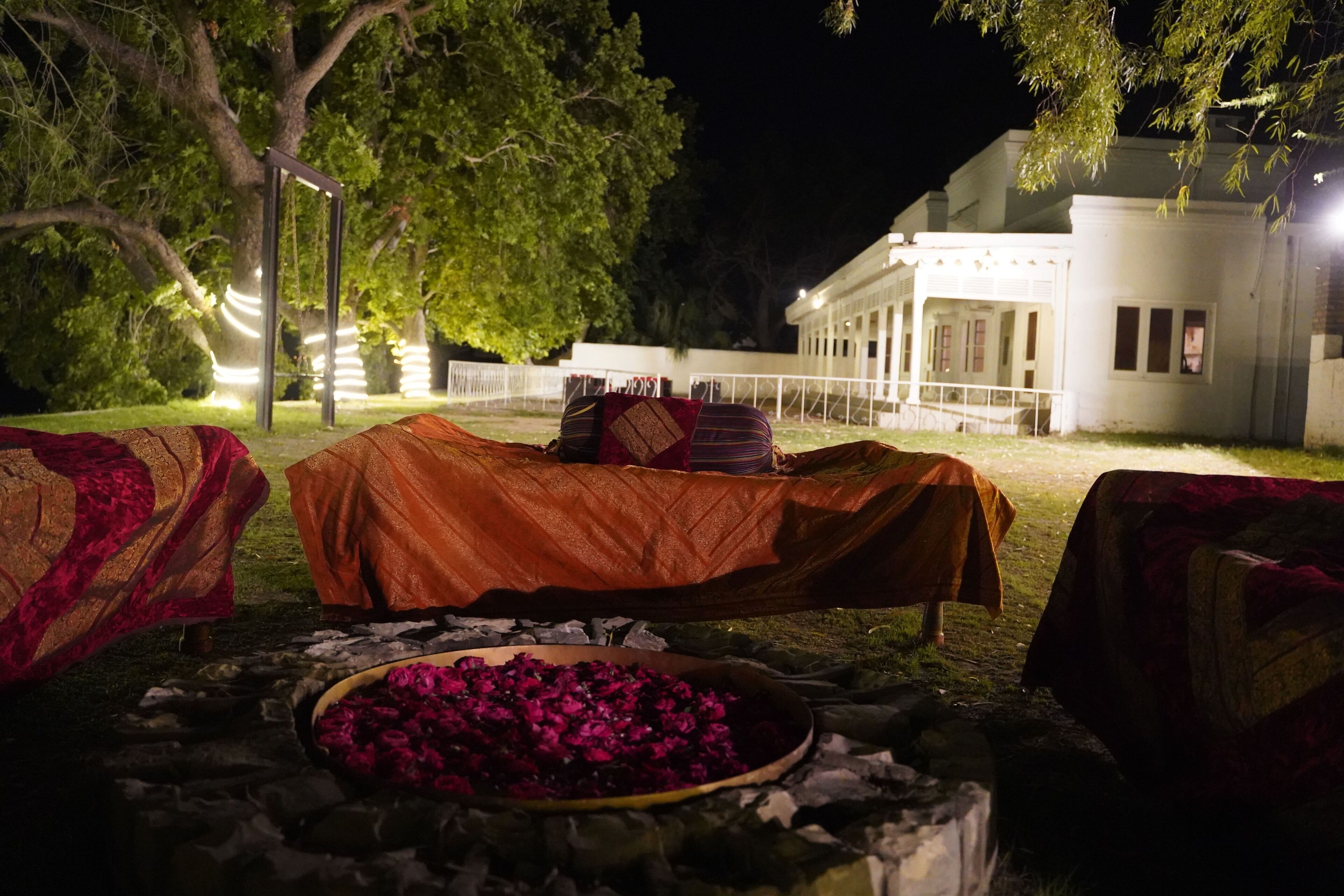 Uma cama com um cobertor vermelho, uma tigela de flores cor-de-rosa e um edifício branco ao fundo.