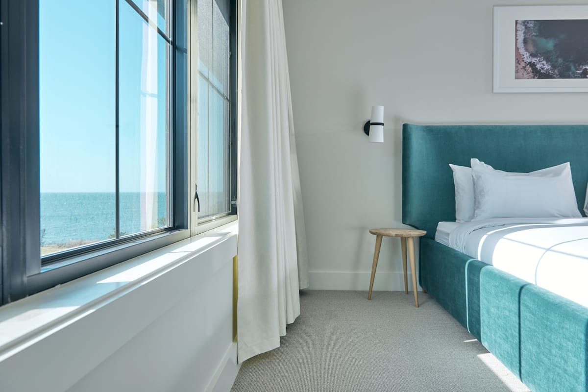 Quarto de hotel com uma grande janela, uma cama com roupa de cama verde-turquesa, uma pequena mesa de cabeceira de madeira e um aplique de parede.