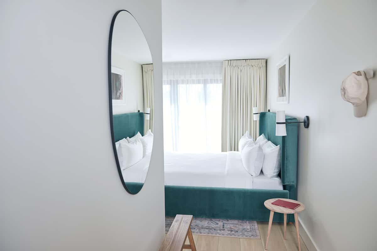 Quarto de hotel com uma cama grande, uma mesa pequena, um espelho e uma janela com cortinas.