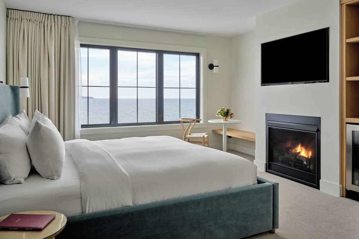 Quarto de hotel com uma cama grande, televisão na parede, lareira e vista para o mar através de uma janela ampla.