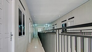 Um corredor longo e estreito com portas e janelas brancas, guarda-corrimão de metal de um lado e chão de azulejos limpo.