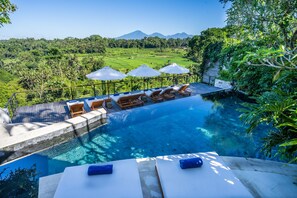 Área de piscina com espreguiçadeiras e guarda-sóis, cercada por vegetação exuberante e vista para montanhas ao longe.