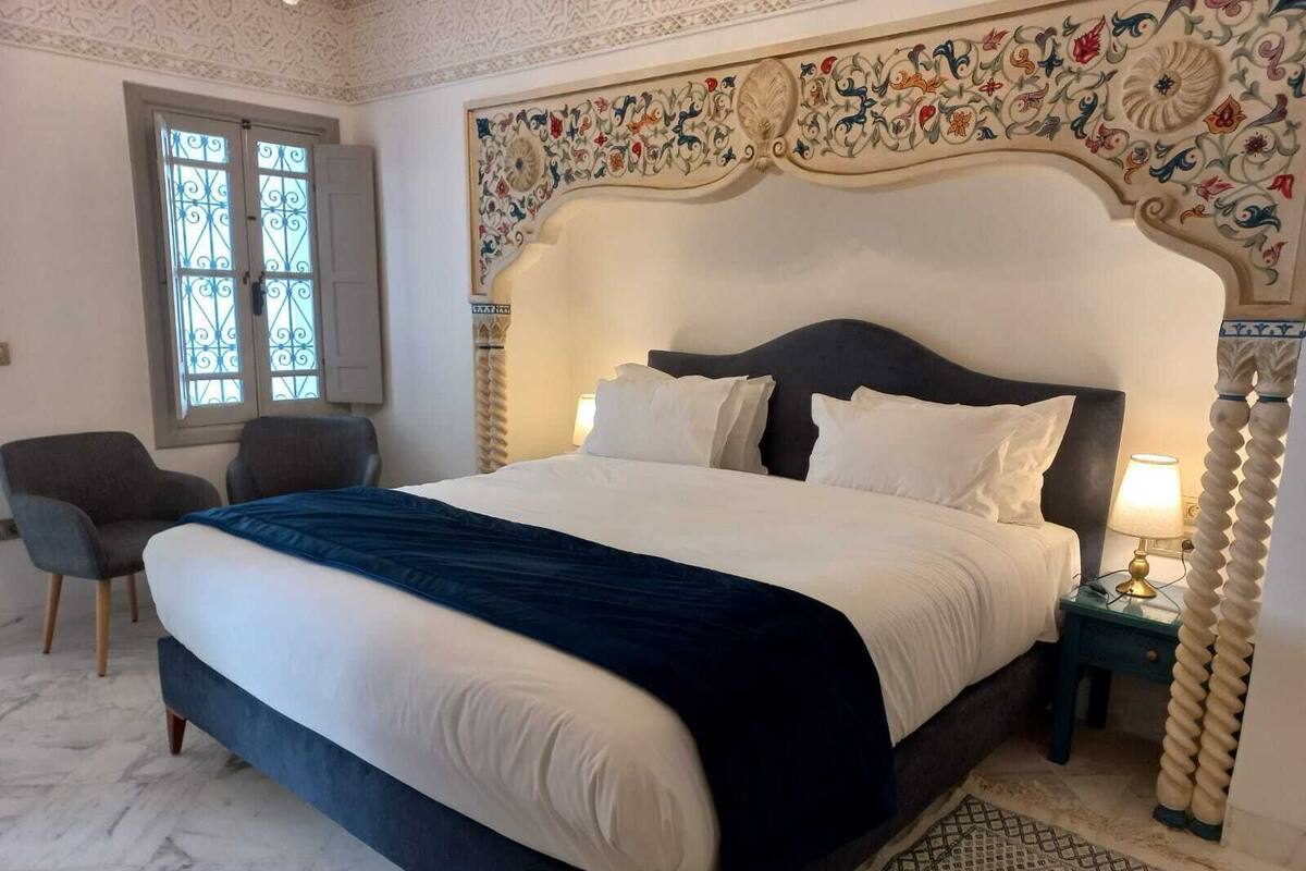 Uma cama grande com cabeceira escura, roupa de cama branca e um painel decorativo na parede. Há uma cadeira e uma mesa de cabeceira com uma luminária.
