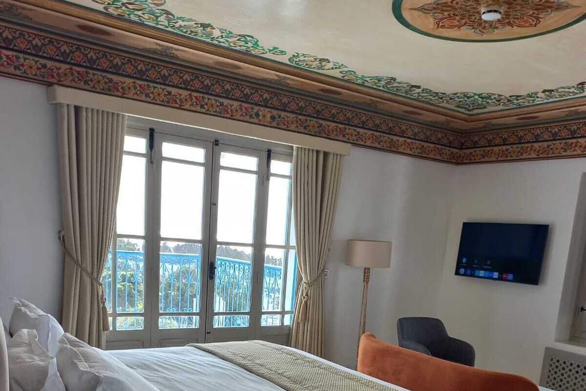 Quarto de hotel com cama, escrivaninha com luminária, televisão e vista para uma varanda.