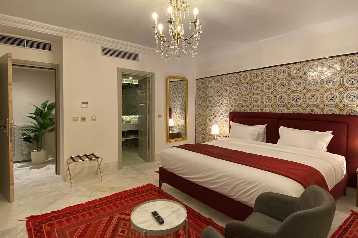 Quarto de hotel com uma cama grande, um candelabro, um tapete vermelho estampado, duas cadeiras cinzas e uma pequena mesa redonda com um controle remoto.