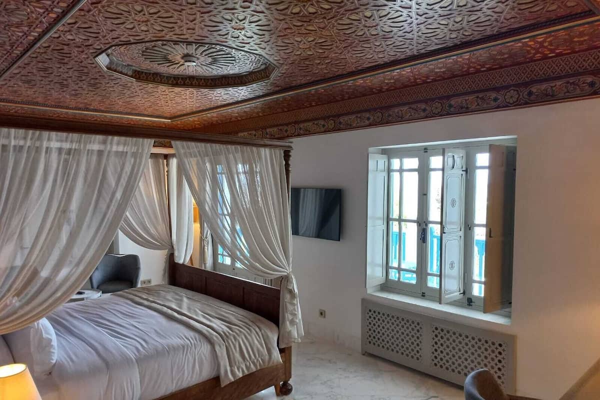 Quarto com cama de dossel, janela com persianas e teto decorativo.
