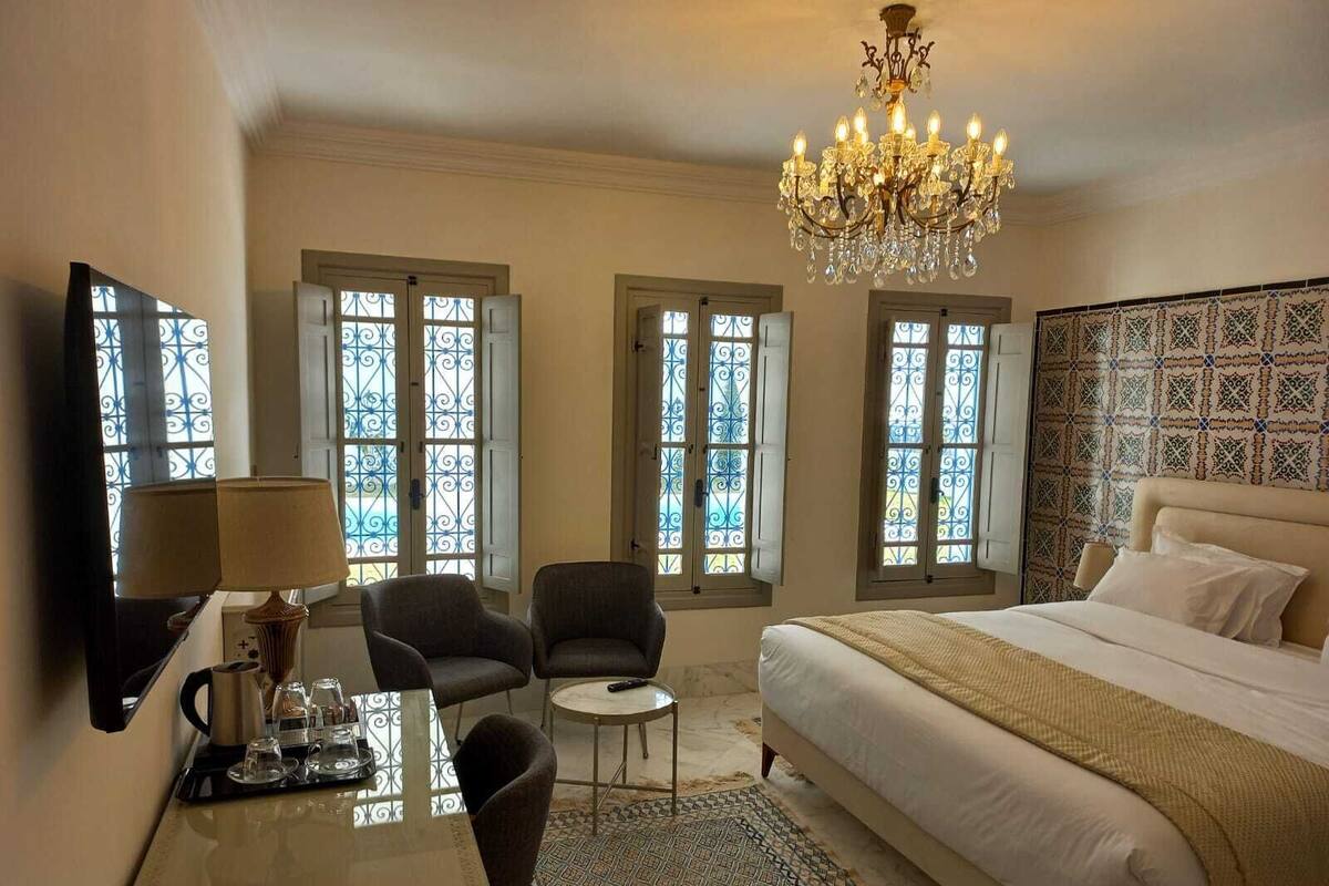 Quarto de hotel com cama, duas poltronas, uma mesinha de centro com abajur, televisão e candelabro.