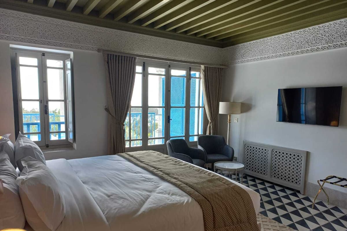 Quarto de hotel com cama, duas poltronas, uma mesa pequena, televisão e varanda com grades azuis.