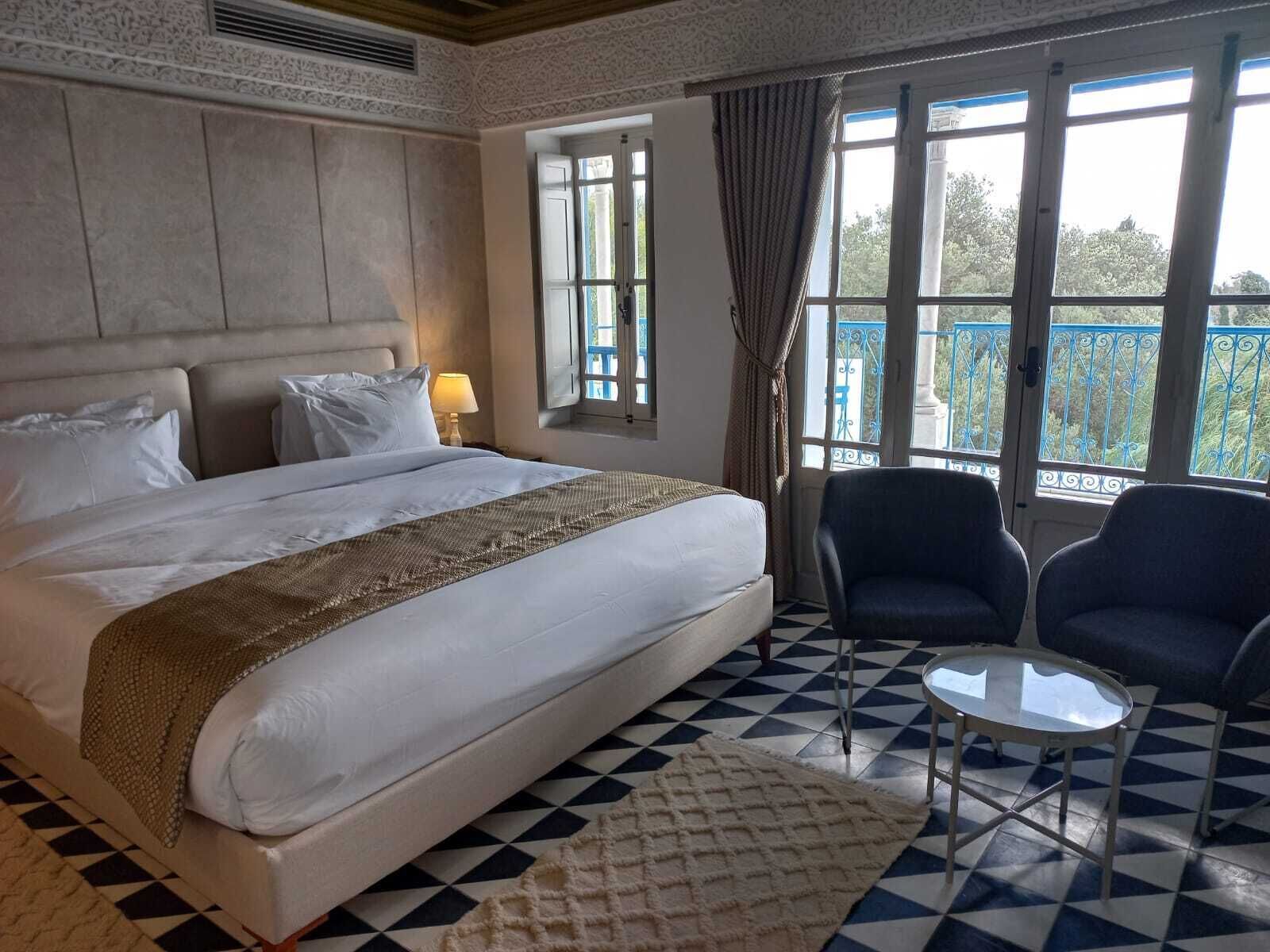 Quarto de hotel com uma cama grande, duas cadeiras, uma mesa pequena e varanda com vista.