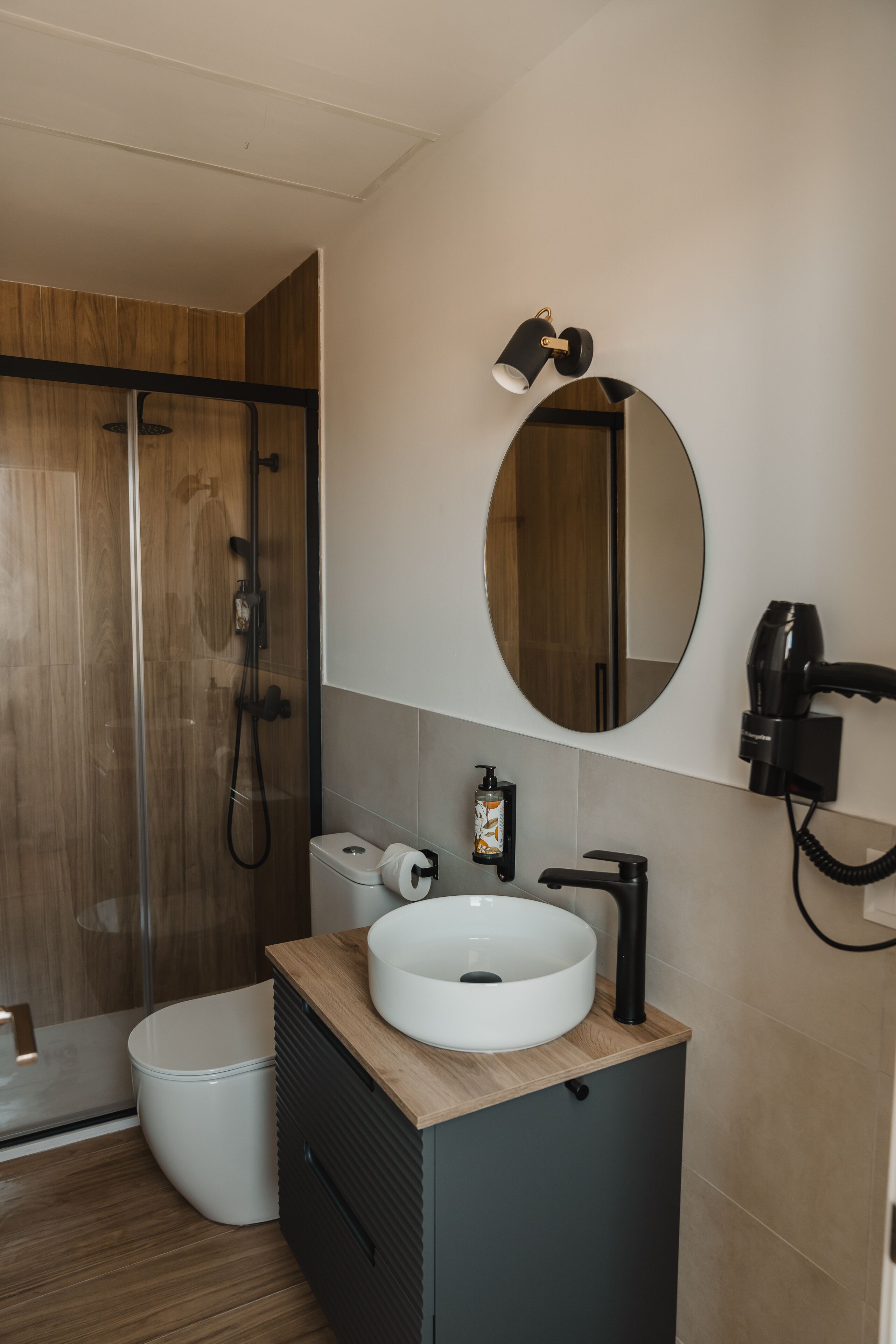 Uma casa de banho moderna com compartimento de duche em madeira, uma sanita branca e um secador de cabelo preto numa prateleira fixada na parede.