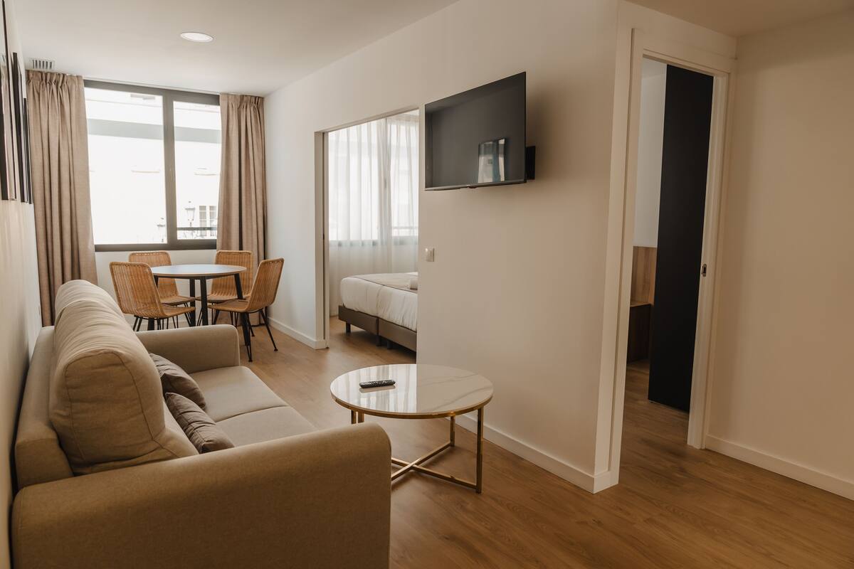 Uma sala de estar moderna com sofá, mesa de jantar e cadeiras, uma TV fixada na parede e uma porta que dá para outro cômodo.