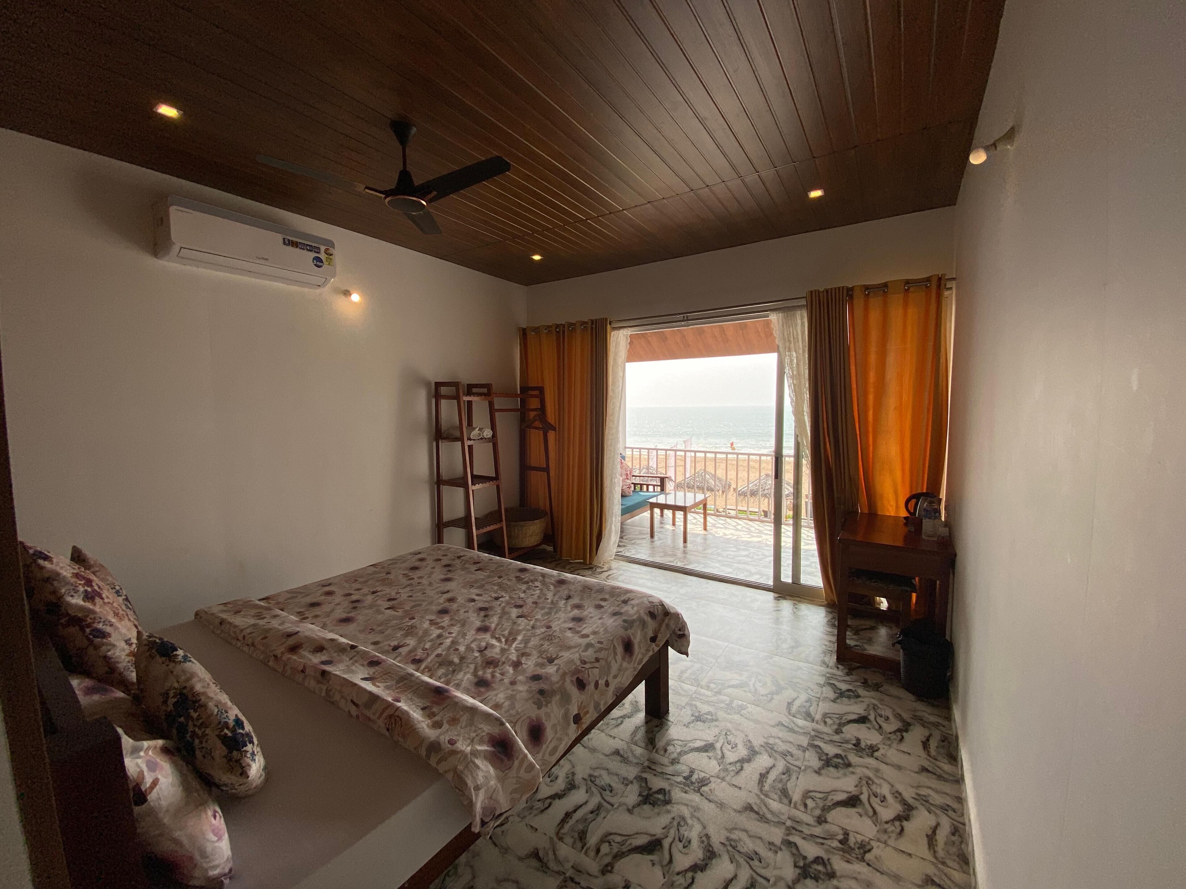 Um quarto com cama, ventilador de teto, ar condicionado, escada de madeira, vista para a praia e varanda com mesa e cadeiras.