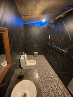 Casa de banho com sanita, lavatório e espelho. As paredes têm um padrão geométrico e existe um candeeiro azul no teto.