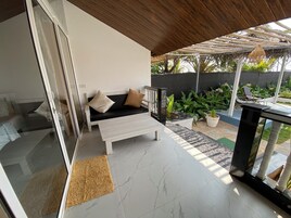 Uma área externa moderna com assento, um banco e almofadas, cercada por plantas em vasos e uma pérgola de madeira.