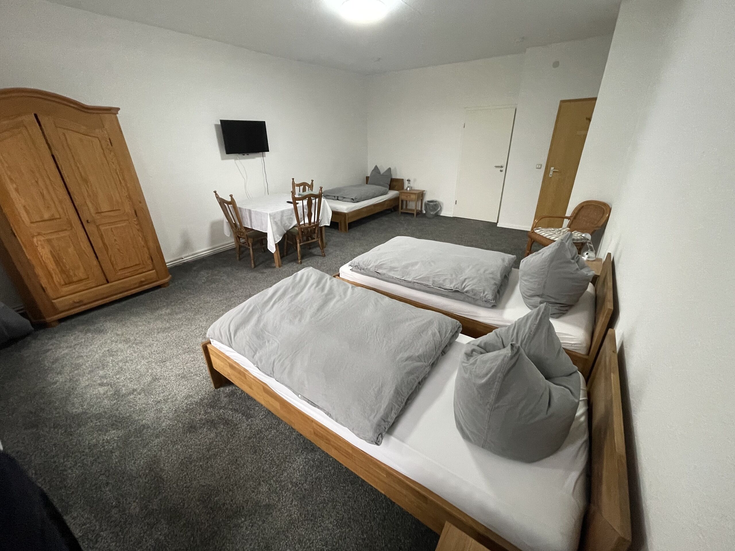 Quarto com duas camas de solteiro, um guarda-roupa de madeira, uma mesa com cadeiras, uma televisão na parede e uma porta.