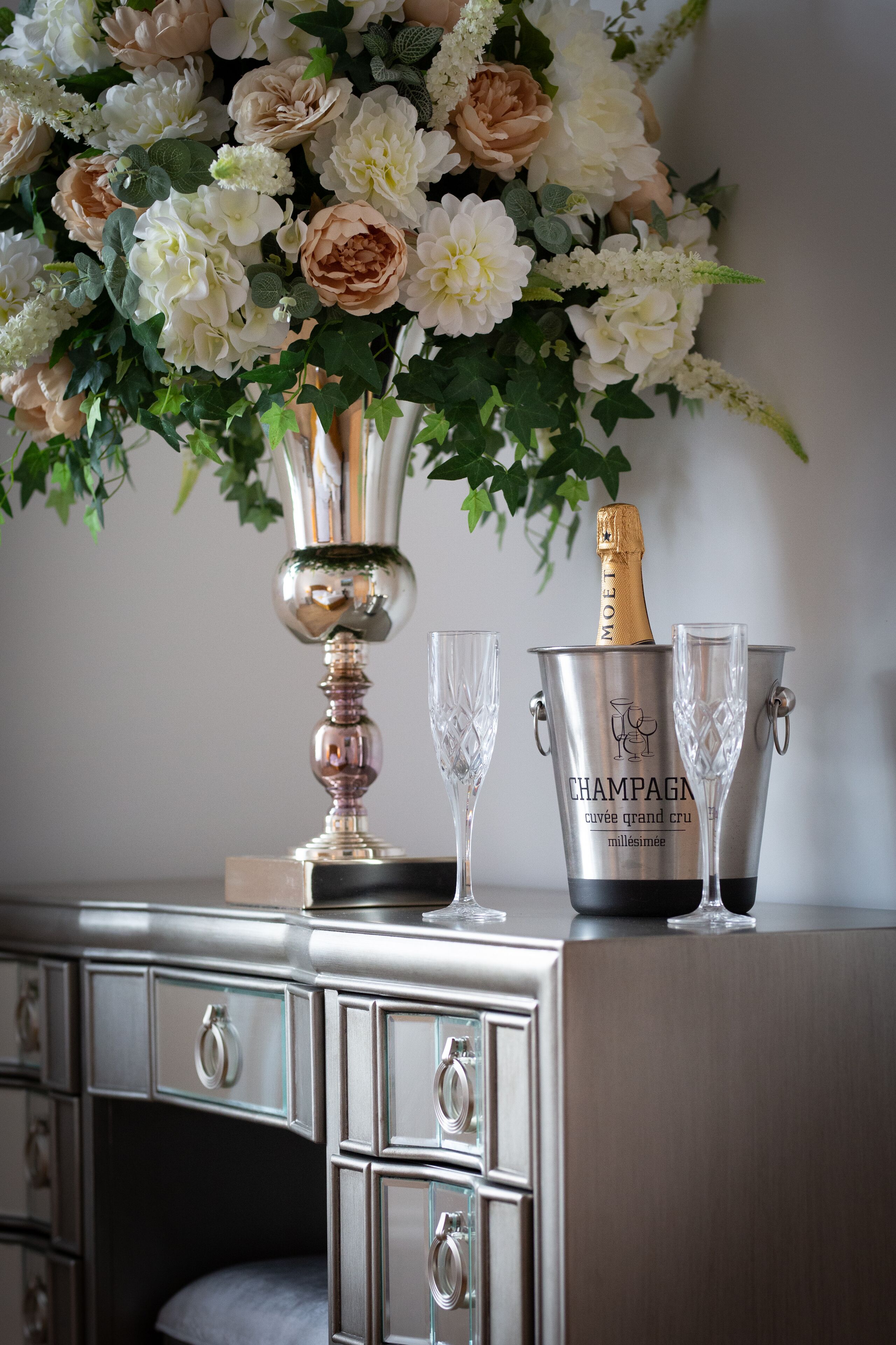 Uma garrafa de champanhe em um balde prateado, com copos ao lado, colocada sobre uma superfície metálica com um vaso decorativo de flores em cima.