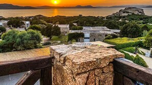 Vista costeira com um pôr do sol, edifícios de pedra e uma formação rochosa imponente.