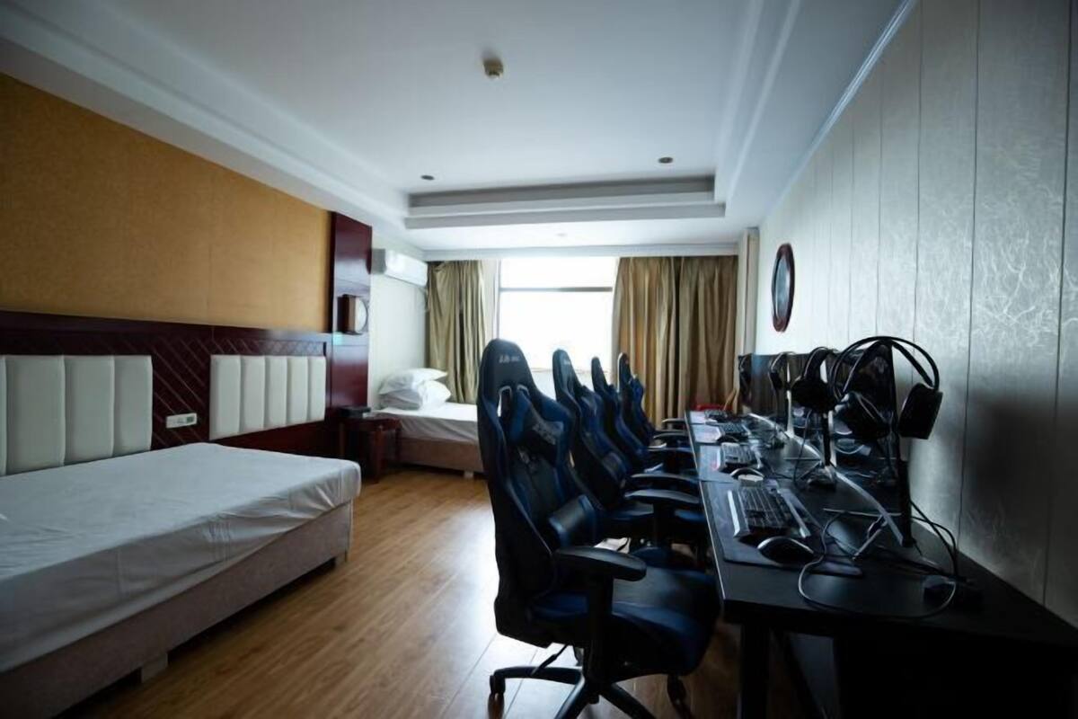 Quarto de hotel com cama, escrivaninha com computador, várias cadeiras de jogos e janela com cortinas.