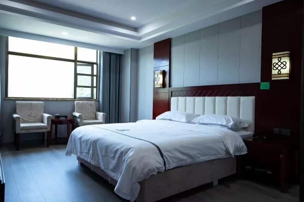 Quarto de hotel com uma cama grande, duas poltronas, uma mesa pequena e uma janela com cortinas.