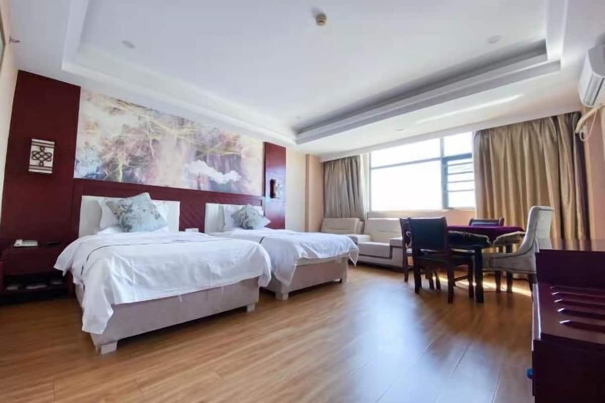 Quarto de hotel com duas camas, uma mesa de jantar, cadeiras, um sofá e uma janela grande com cortinas.