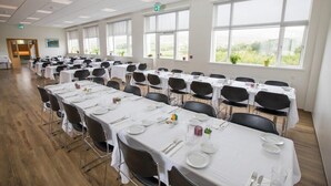 Sala de conferências com várias mesas preparadas para refeições, grandes janelas e vista para o verde lá fora.