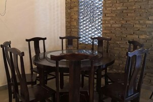 Mesa de jantar redonda de madeira com seis cadeiras, apoiada em parede de tijolos com janela decorada.