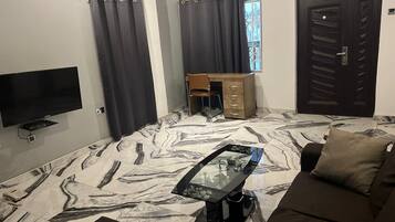 Uma sala de estar com televisão de tela plana, uma mesa de centro de vidro, um sofá, uma escrivaninha de madeira e uma porta escura.