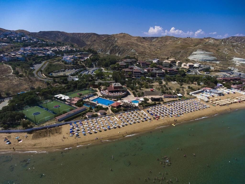 Vista aérea de um resort à beira-mar, com praia, piscinas e montanhas ao fundo.