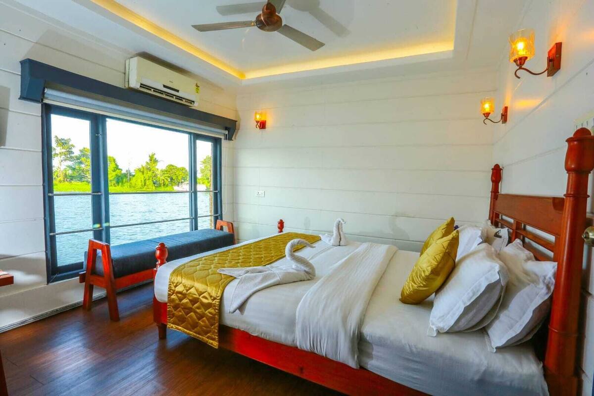 Quarto de hotel com uma cama grande, janela vista para a água, ventilador de teto e luminárias na parede.