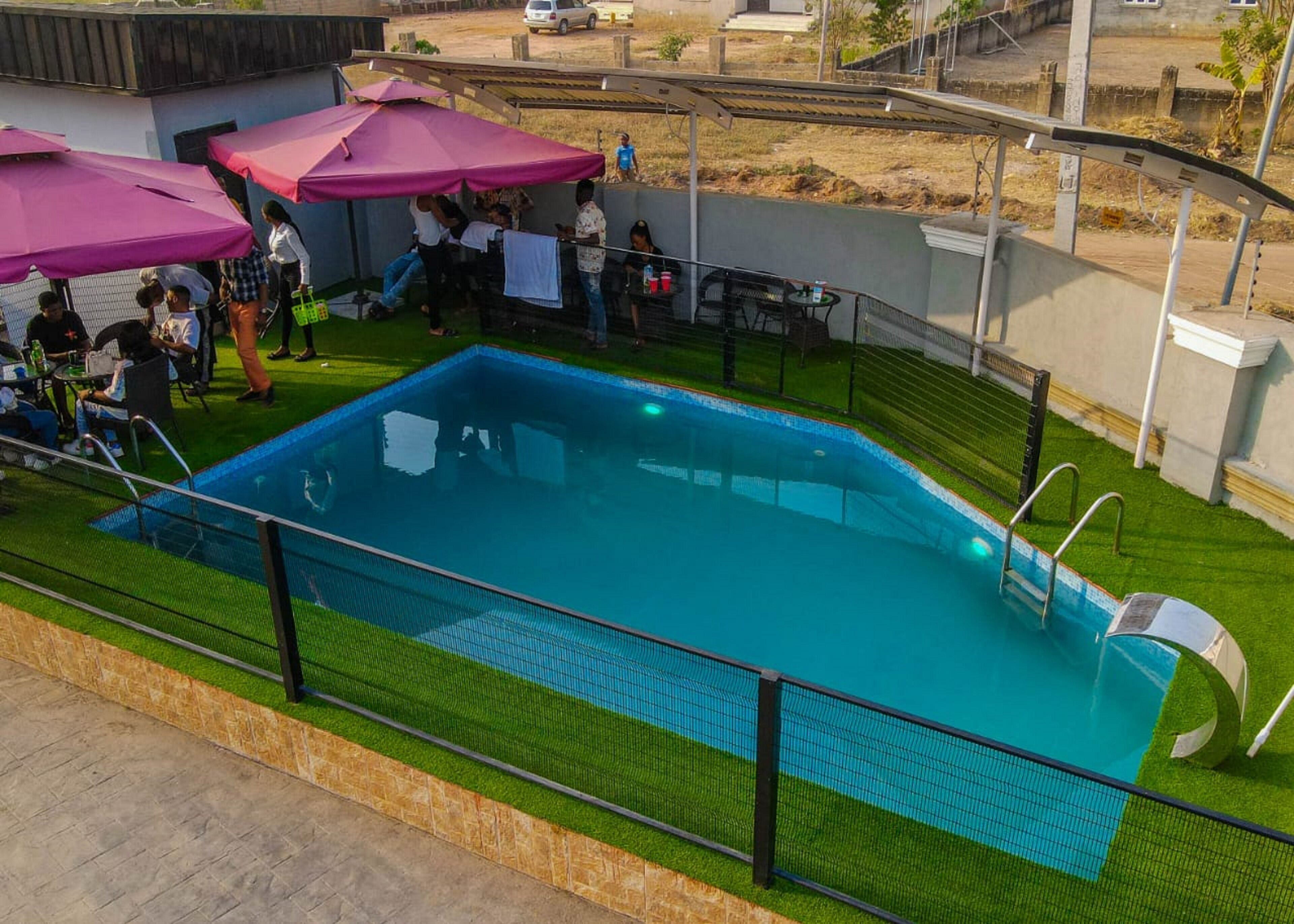 Uma piscina retangular com grade de proteção, rodeada por grama sintética, e protegida por grandes guarda-sóis.