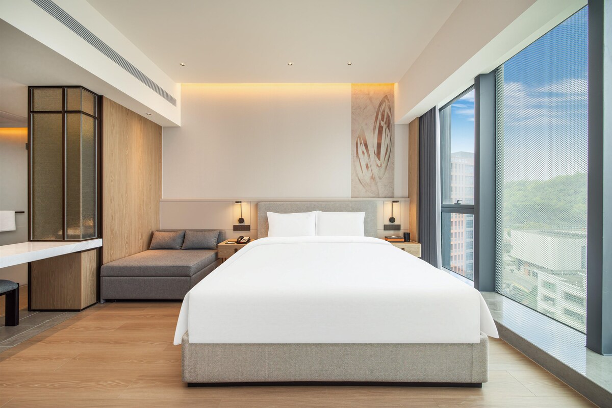 Quarto de hotel moderno com uma cama grande, área de estar, escrivaninha e uma janela ampla com vista para a cidade.