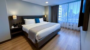 Quarto de hotel moderno com uma cama grande, abajures, mesa de cabeceira e vista da cidade através de cortinas leves.