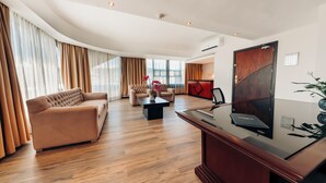 Quarto de hotel moderno com piso de madeira, um sofá, uma poltrona, uma escrivaninha com computador e uma televisão grande.