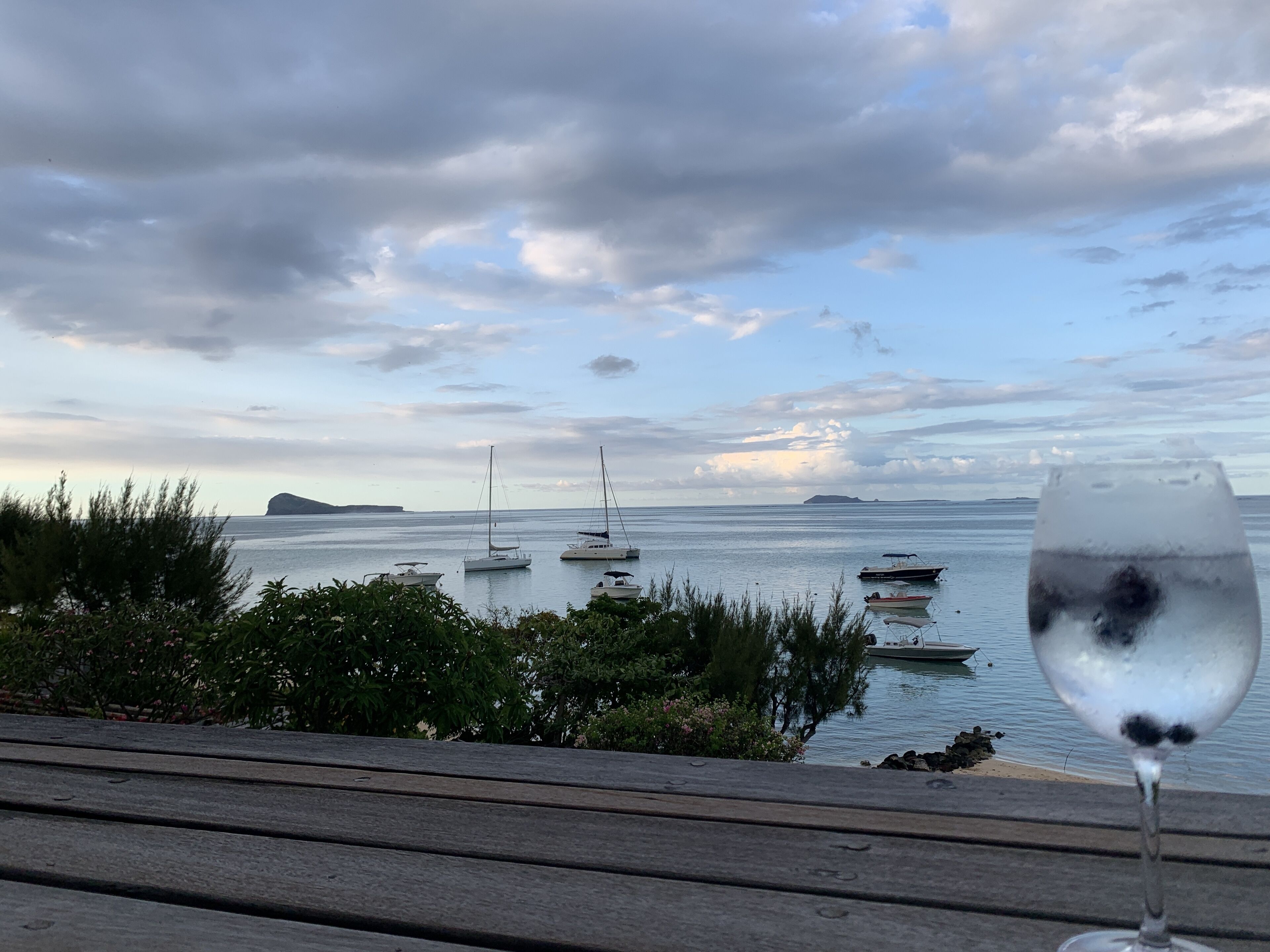 Um copo de água sobre um banco de madeira com vista para um mar calmo, barcos e ilhas ao longe.