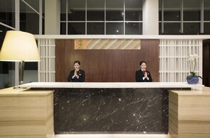 Balcão de recepção com dois funcionários, bancada de mármore, luminária e painel decorativo na parede.