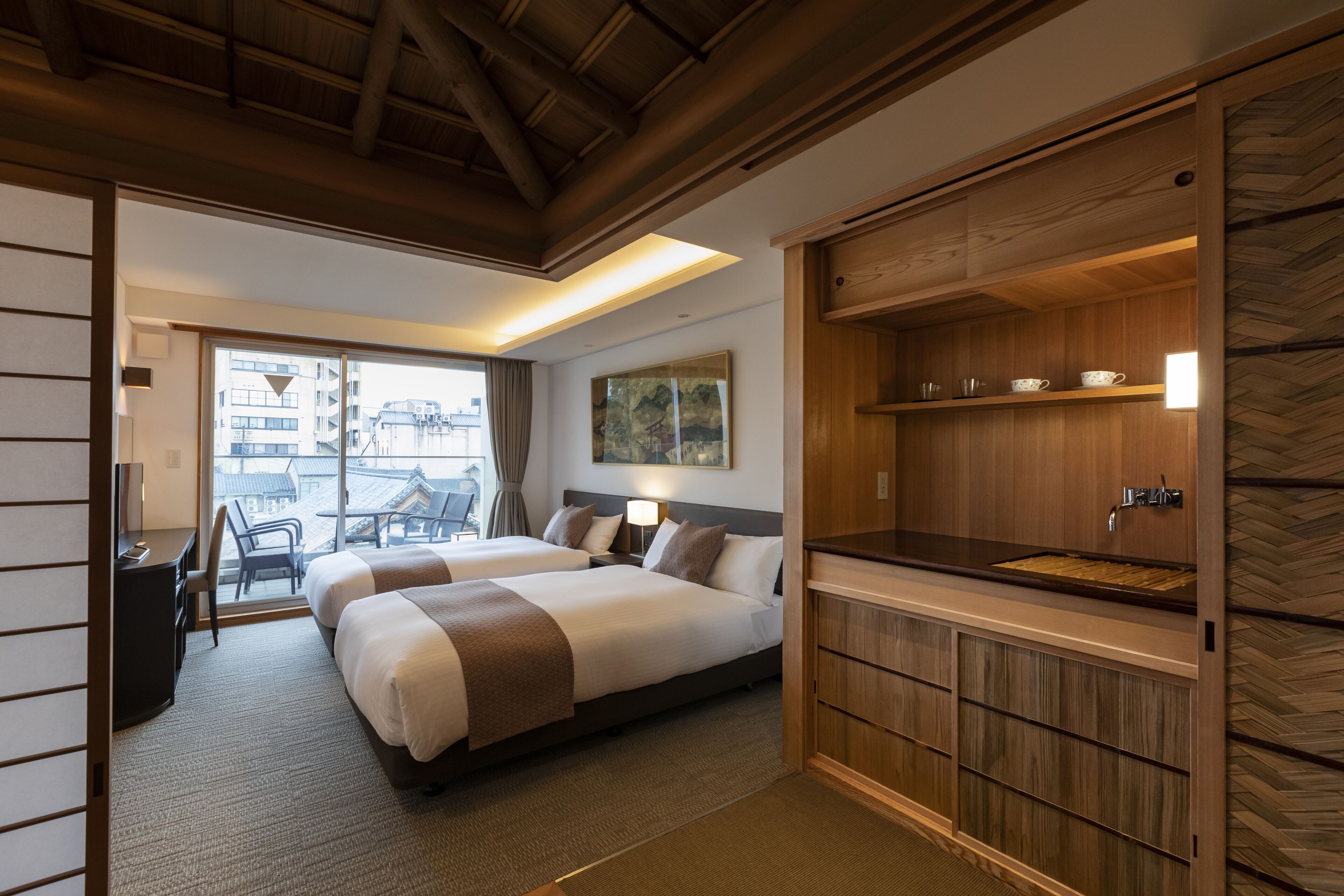 Quarto de hotel com duas camas, uma escrivaninha, uma cadeira, uma janela grande e um armário de madeira com uma pia.